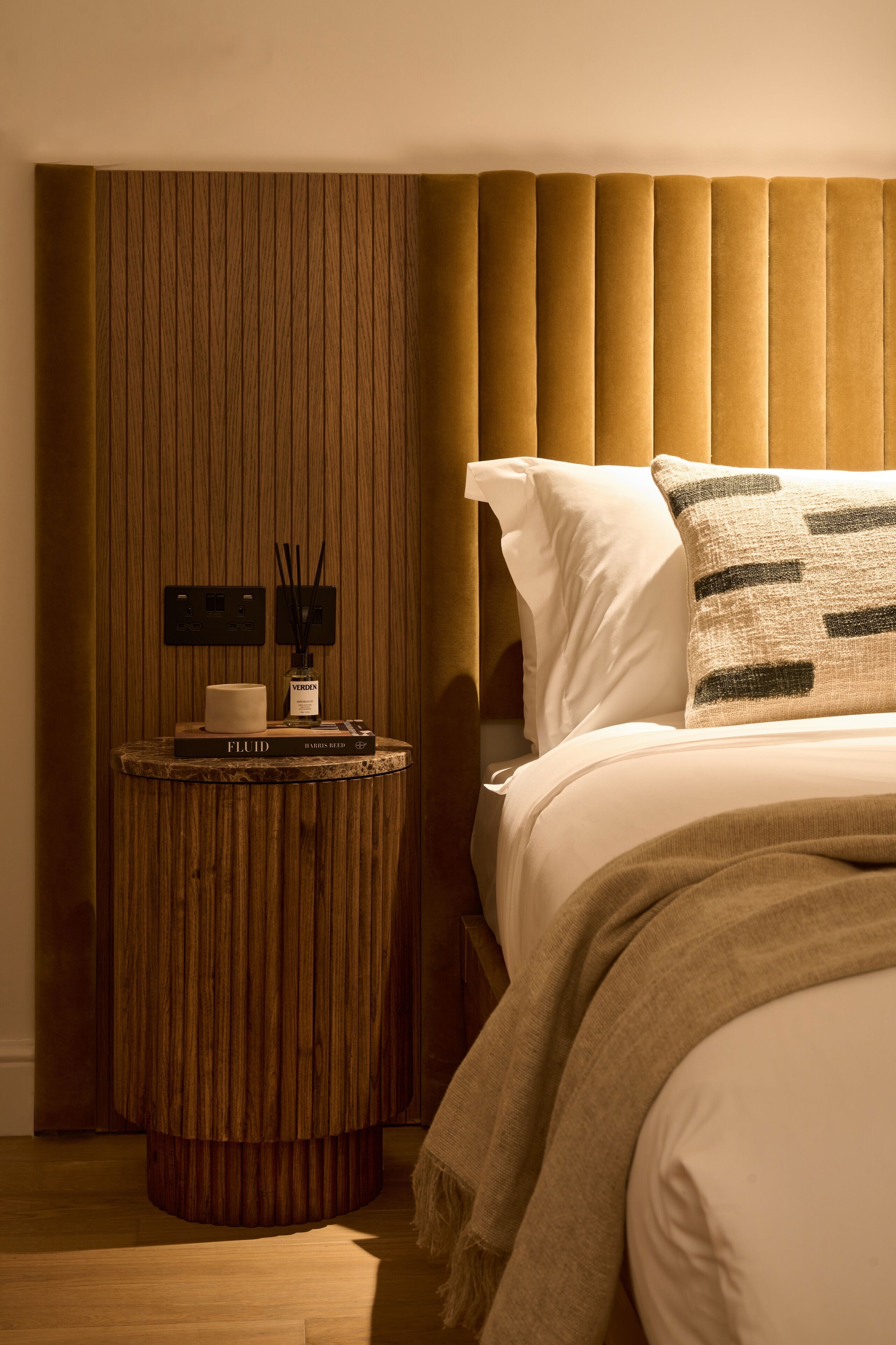 Quarto com cabeceira de madeira, mesa de cabeceira com uma garrafa e controle remoto, e cama com cobertor e almofadas bege.