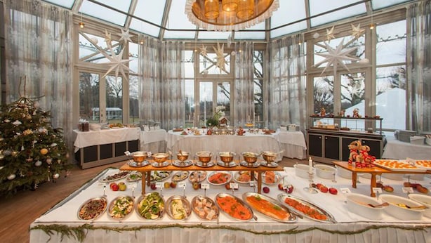 Um quarto amplo com teto alto e grandes janelas, com um buffet repleto de diversos alimentos e uma árvore de Natal.