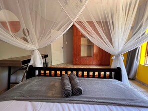 Cama com manta cinza e duas toalhas enroladas, cabeceira de madeira e dossel branco.