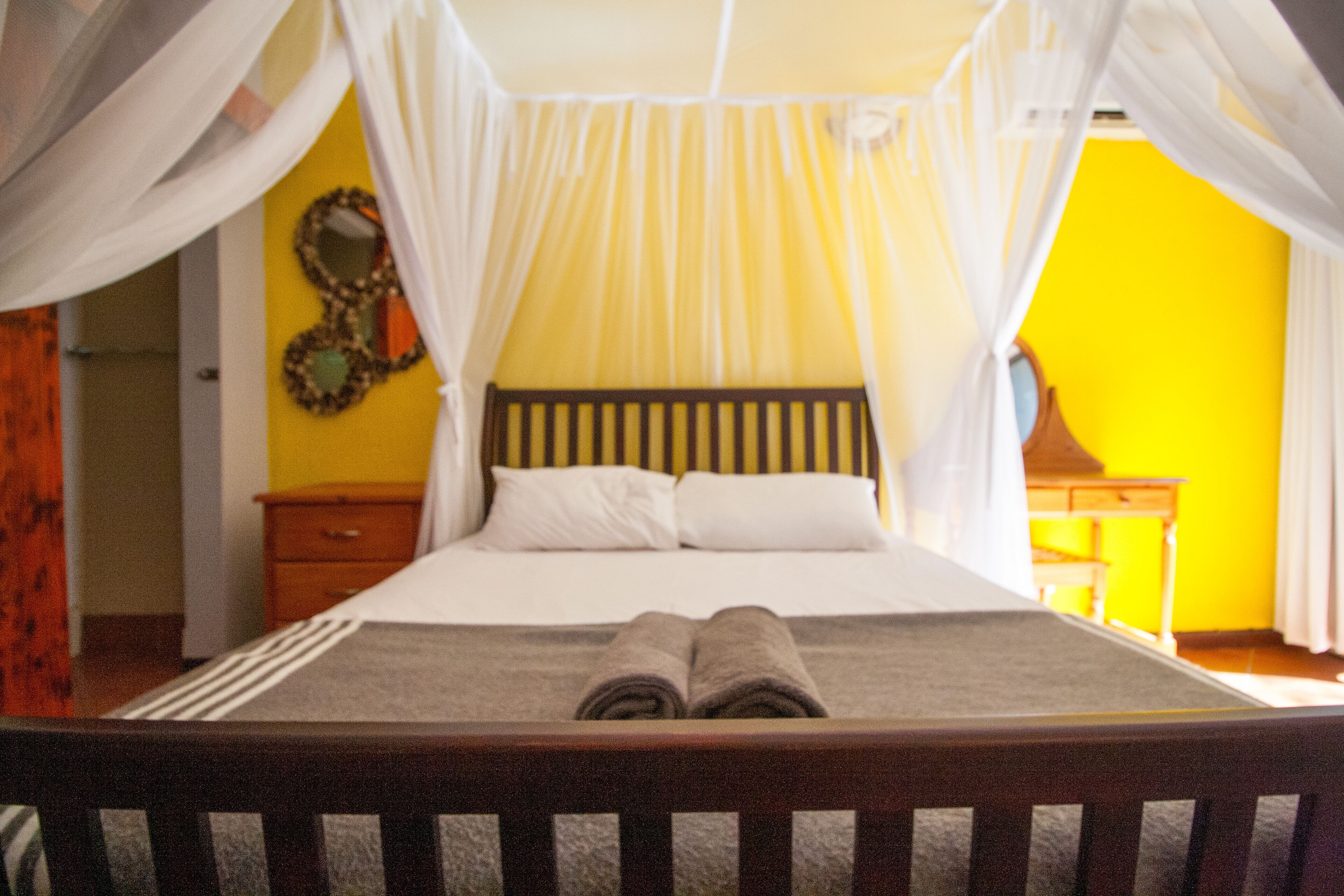 Cama com roupa de cama branca e toalha enrolada, cabeceira de madeira e parede amarela com espelho.