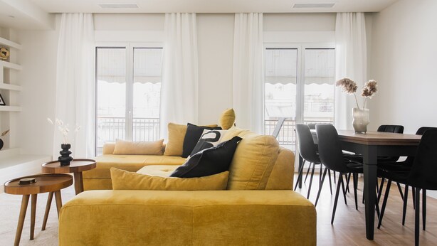 Uma sala de estar moderna com um sofá amarelo, uma mesa de jantar com cadeiras e grandes janelas com cortinas brancas.