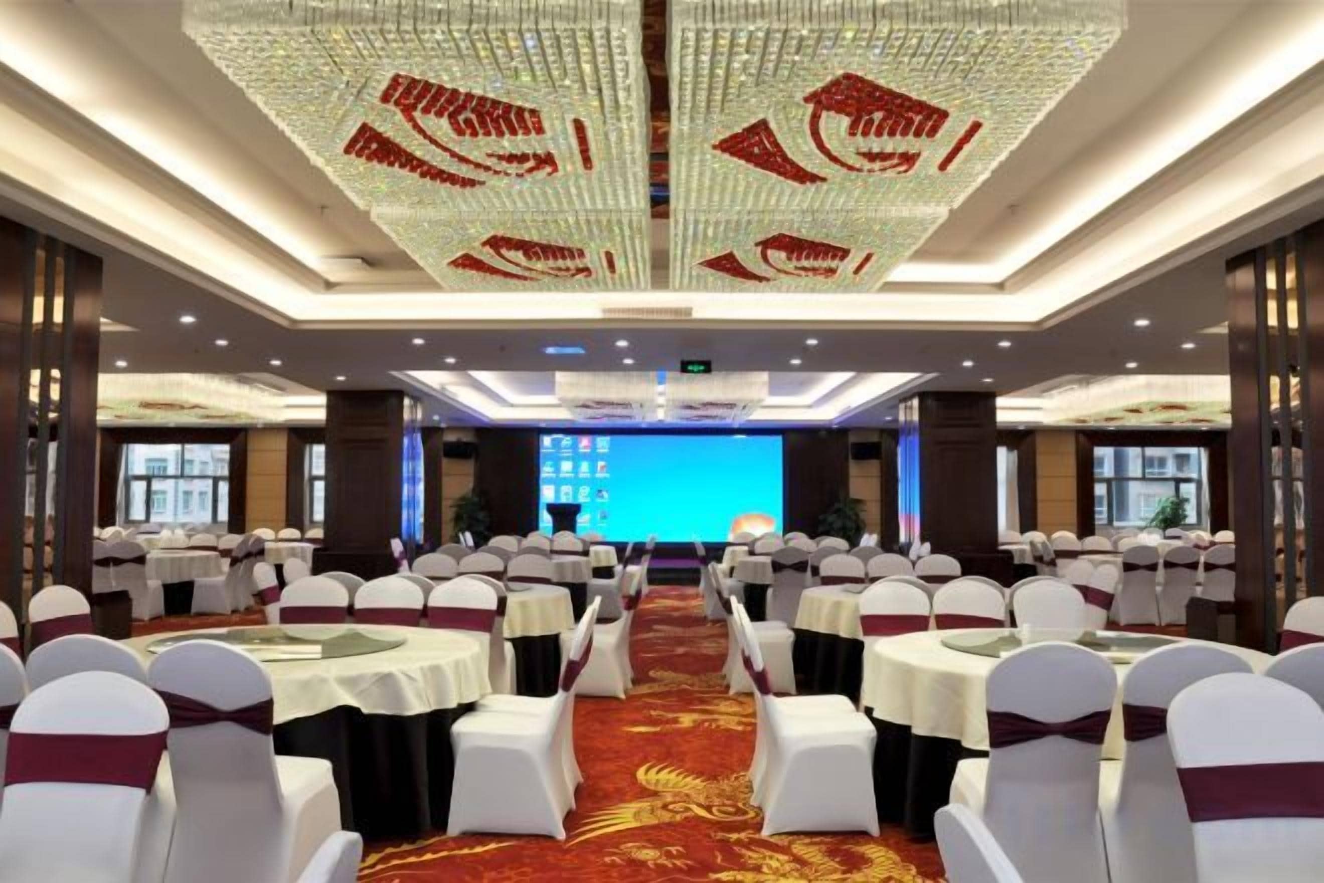 Uma sala de banquetes com fileiras de mesas e cadeiras, um palco com uma tela, e um tapete vermelho.