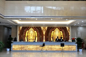 Área de recepção de hotel com balcão de mármore, dois recepcionistas e um grande painel decorativo.