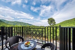 Varanda com cadeiras de vime e uma mesa, uma fruteira e vista para colinas verdes sob um céu parcialmente nublado.