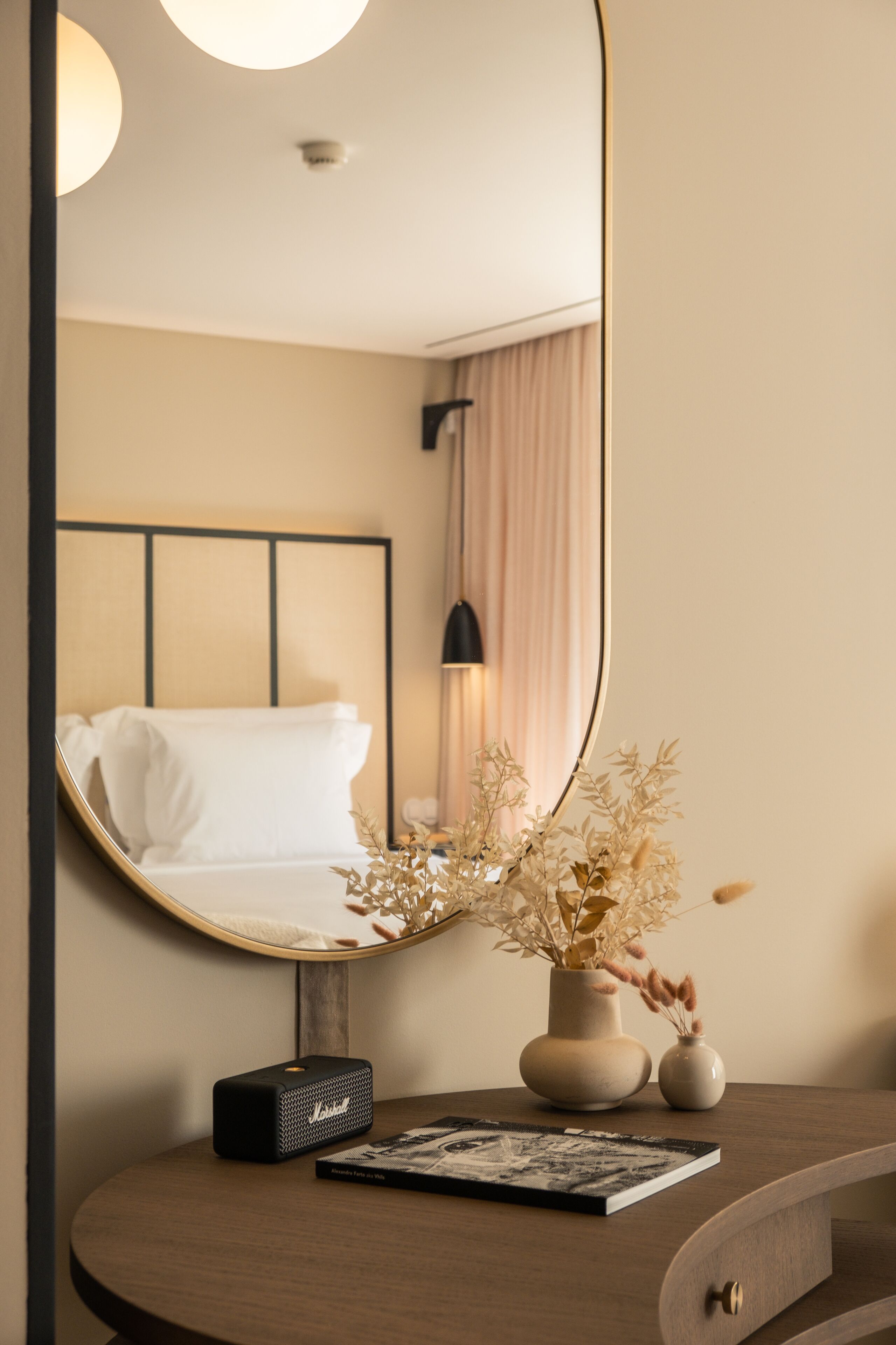 Quarto de hotel com cama, mesa de cabeceira com luminária, espelho e um vaso com flores secas.