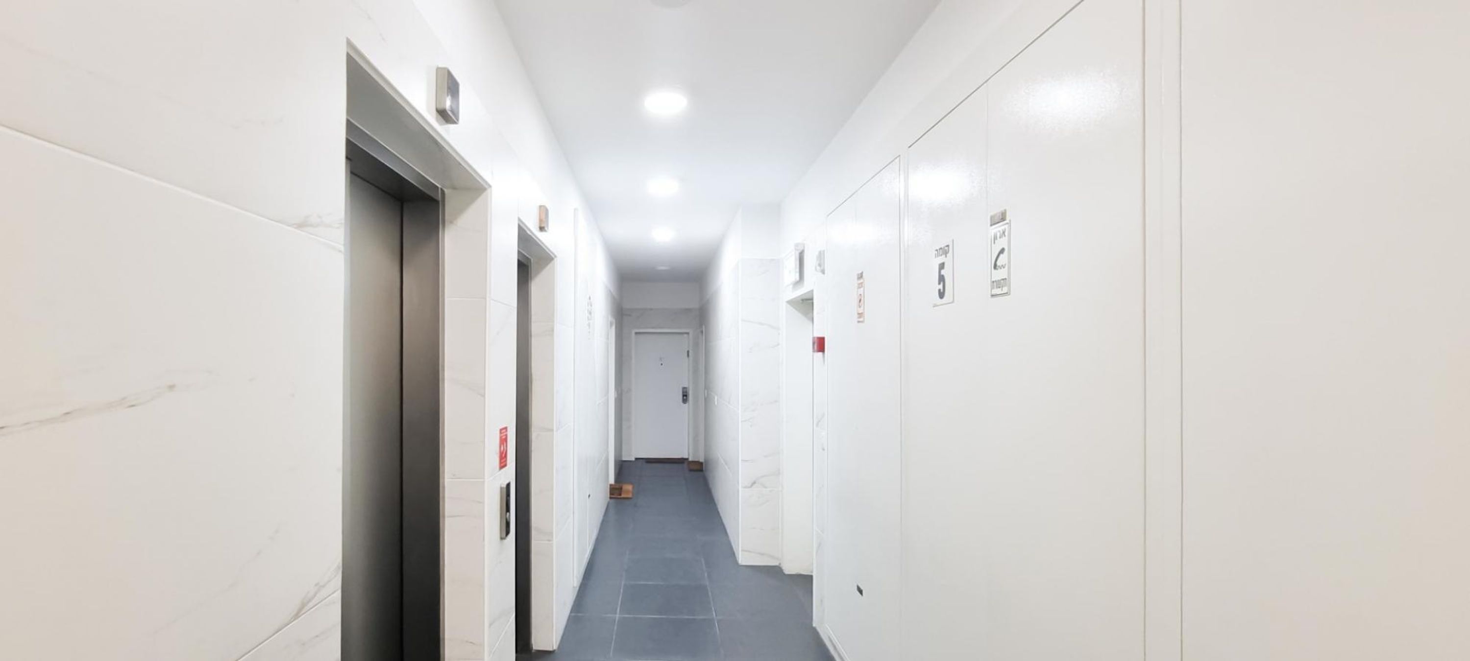 Um corredor estreito e bem iluminado, com paredes brancas e chão de azulejos, que termina numa porta.