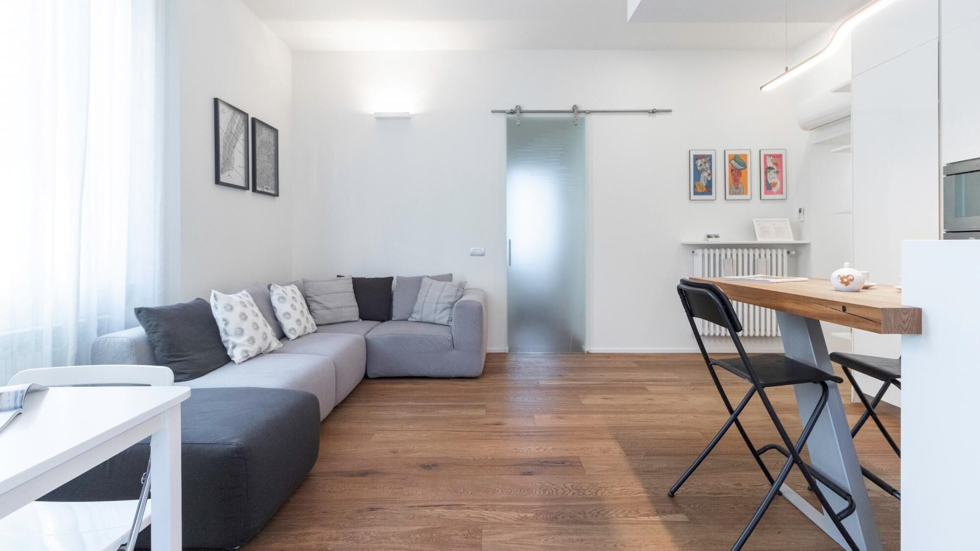 Uma sala de estar moderna com um sofá seccional cinza, uma mesa de jantar de madeira com cadeiras e uma área de cozinha com radiador e obras de arte emolduradas na parede.