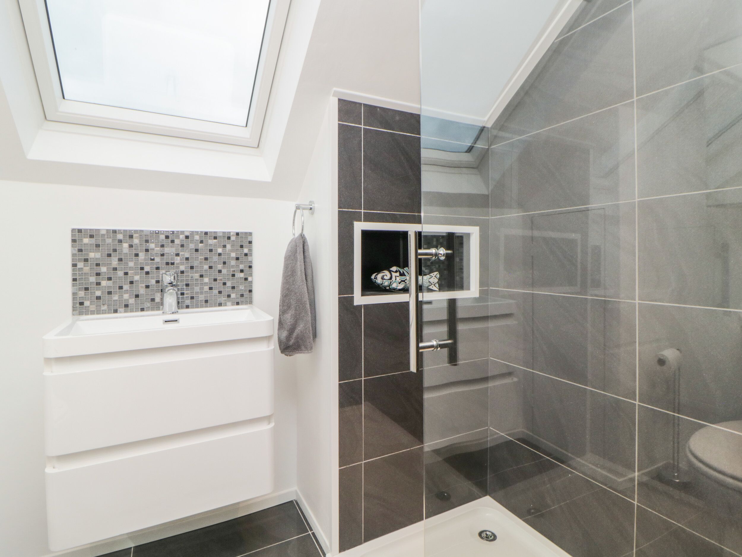 Um banheiro moderno com claraboia, chuveiro com box de vidro e toucador branco com revestimento de azulejos.