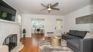 Uma sala de estar com lareira, um sofá cinza, um ventilador de teto e uma janela grande.