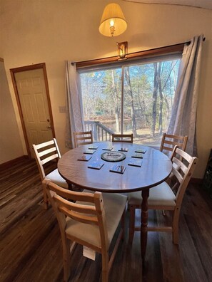 Mesa de jantar redonda de madeira com seis cadeiras, uma luminária acima e vista para árvores pela janela.