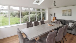 Uma sala de jantar com uma mesa comprida e cadeiras, janelas amplas, um violão e um sofá.