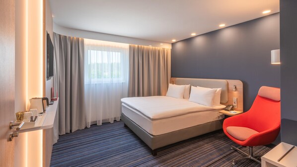 Quarto de hotel moderno com uma cama grande, uma cadeira vermelha, uma mesa de cabeceira e uma janela com cortinas.