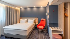Quarto de hotel com cama, uma cadeira vermelha, uma escrivaninha pequena e um guarda-roupa.