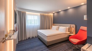 Quarto de hotel moderno com uma cama grande, uma cadeira vermelha, uma mesa de cabeceira e uma janela com cortinas.