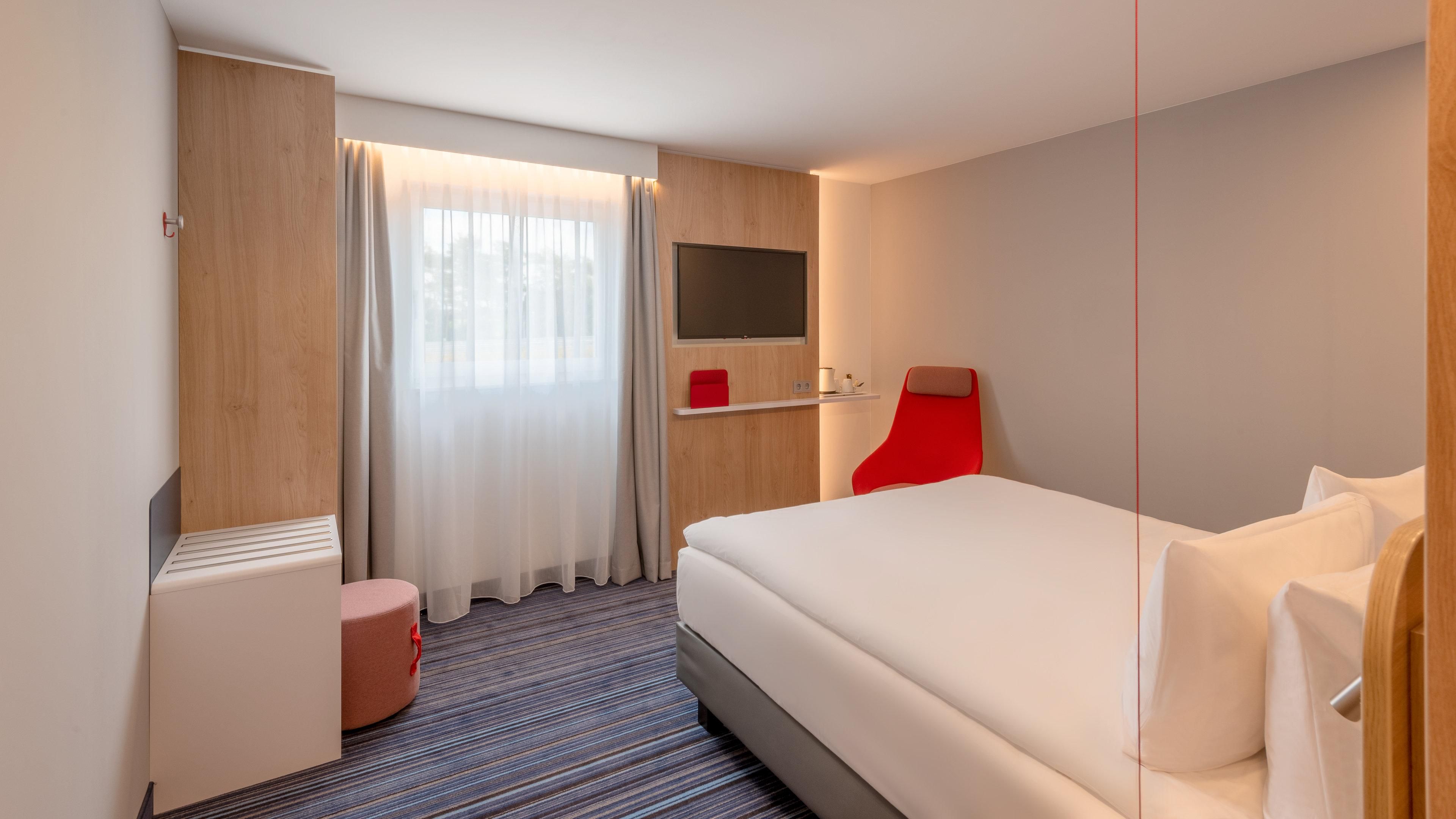 Quarto de hotel moderno com uma cama grande, televisão de tela plana na parede, uma cadeira vermelha e uma mesinha com um almofado vermelho.