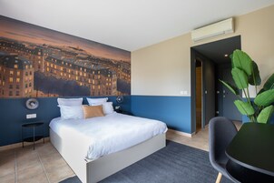 Quarto de hotel com uma cama grande, uma mesa pequena, uma cadeira e uma janela ampla com vista para a cidade.