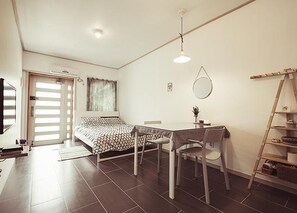 Quarto compacto com cama, escrivaninha e cadeira. Há uma janela com cortinas, uma porta e um pequeno prateleira com objetos.