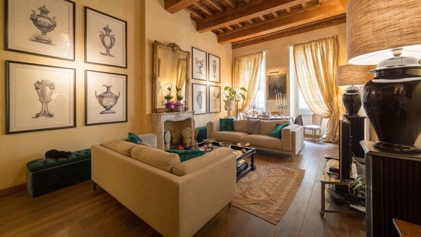 Uma sala de estar com sofá, poltronas, mesa de centro e lareira. Há quadros nas paredes e um abajur sobre uma mesa.