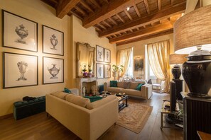 Uma sala de estar com sofá, poltronas, mesa de centro e lareira. Há quadros nas paredes e um abajur sobre uma mesa.