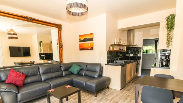 Uma sala moderna com sofá seccional, uma mesa de centro e um espaço de jantar com mesa e cadeiras. Uma cozinha com bancada e eletrodomésticos é visível ao fundo.