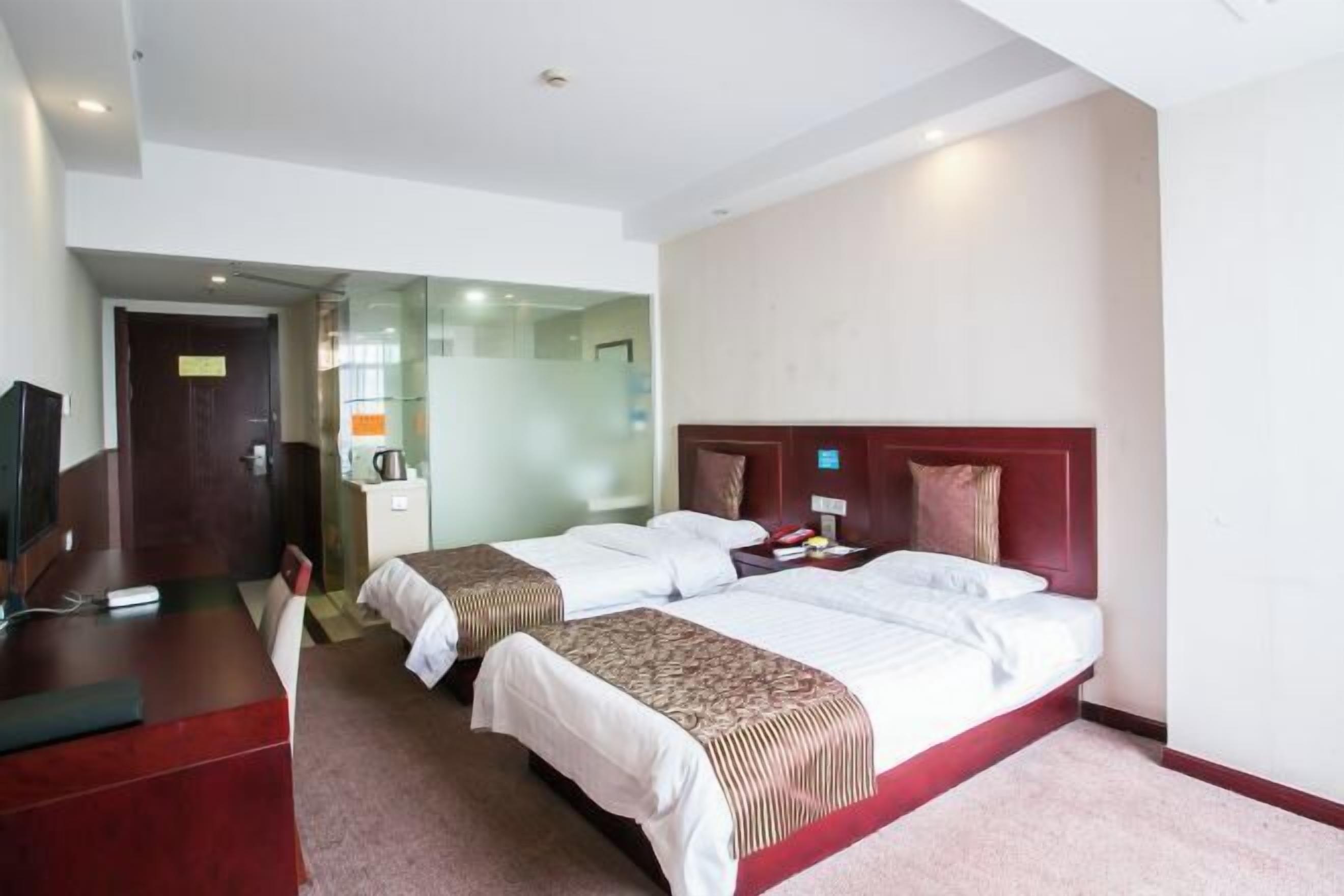 Quarto de hotel com duas camas, uma mesa de escritório, televisão e banheiro visível através de um painel de vidro.