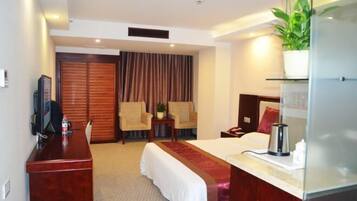 Quarto de hotel com cama, escrivaninha, duas cadeiras, televisão, planta e janela com cortinas.