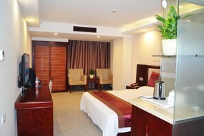 Quarto de hotel com cama, escrivaninha, duas cadeiras, televisão, planta e janela com cortinas.