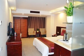 Quarto de hotel com cama, escrivaninha, cadeiras, televisão, planta e janela.