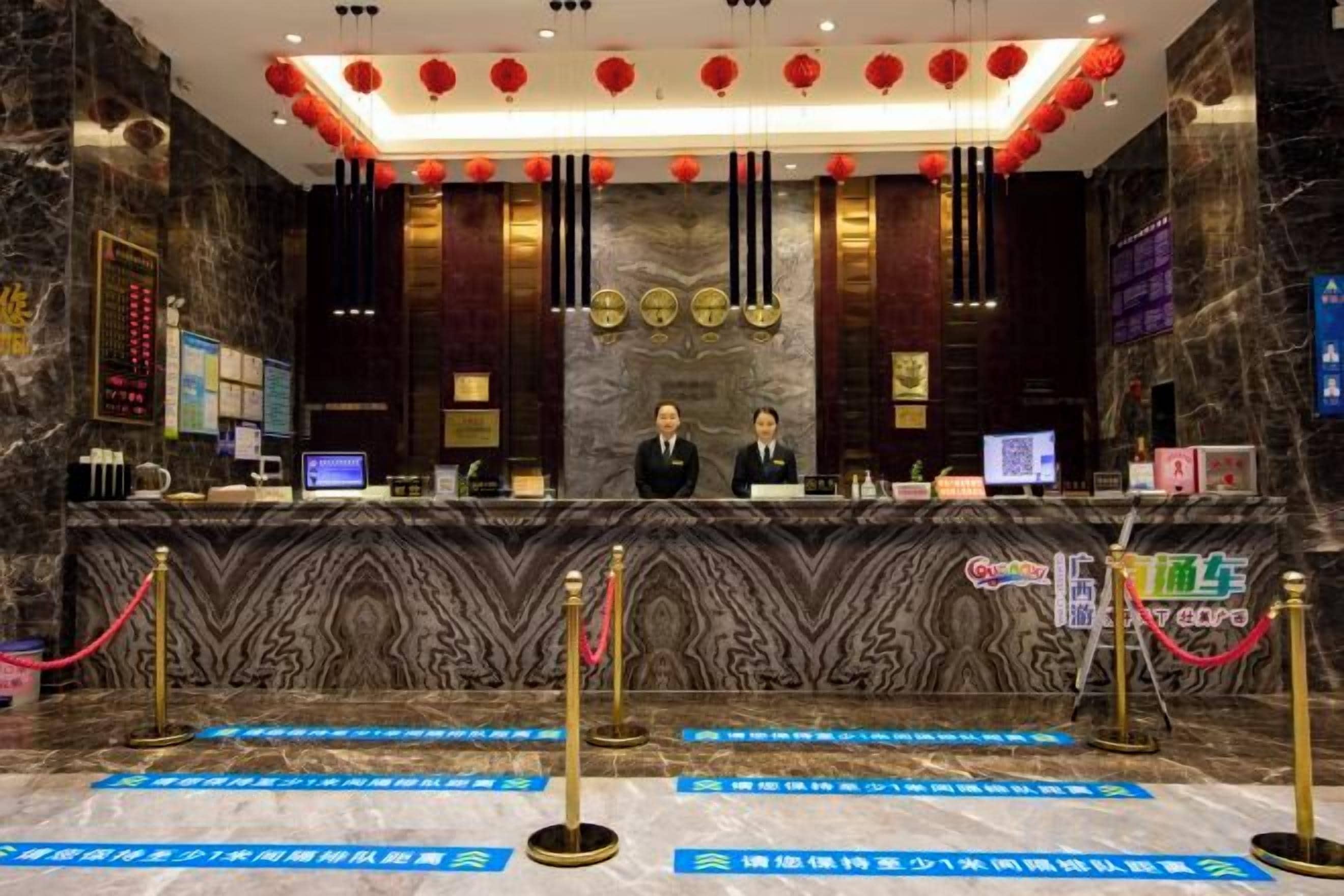 Área de recepção de hotel com balcão de mármore, mesa de atendimento e dois recepcionistas.