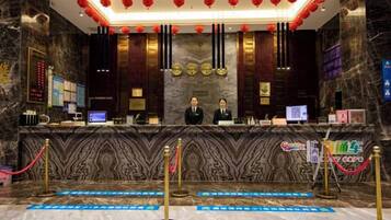 Área de recepção de hotel com balcão de mármore, mesa de atendimento e dois recepcionistas.