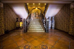 Uma escadaria imponente com corrimãos decorados, que leva a um espaço com paredes estampadas e piso de azulejos.
