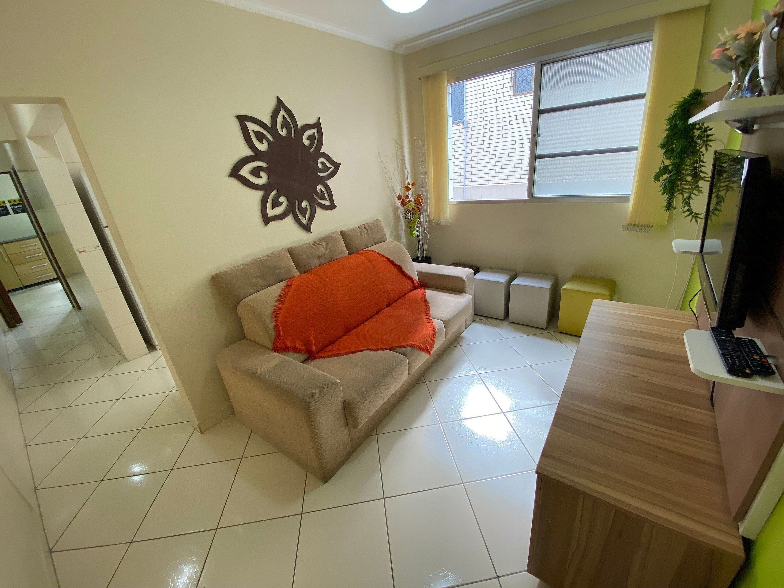Uma sala de estar com um sofá bege, um cobertor laranja, um objeto decorativo na parede, uma janela com persianas e uma TV sobre um móvel.