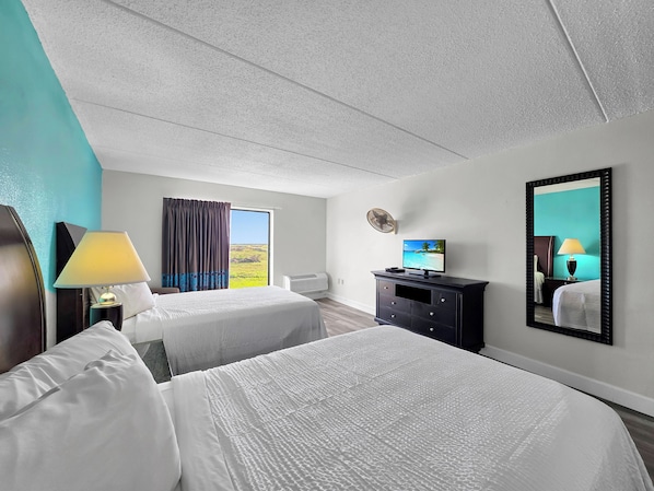 Quarto de hotel com duas camas, televisão, escrivaninha, espelho e janela com vista para a praia.