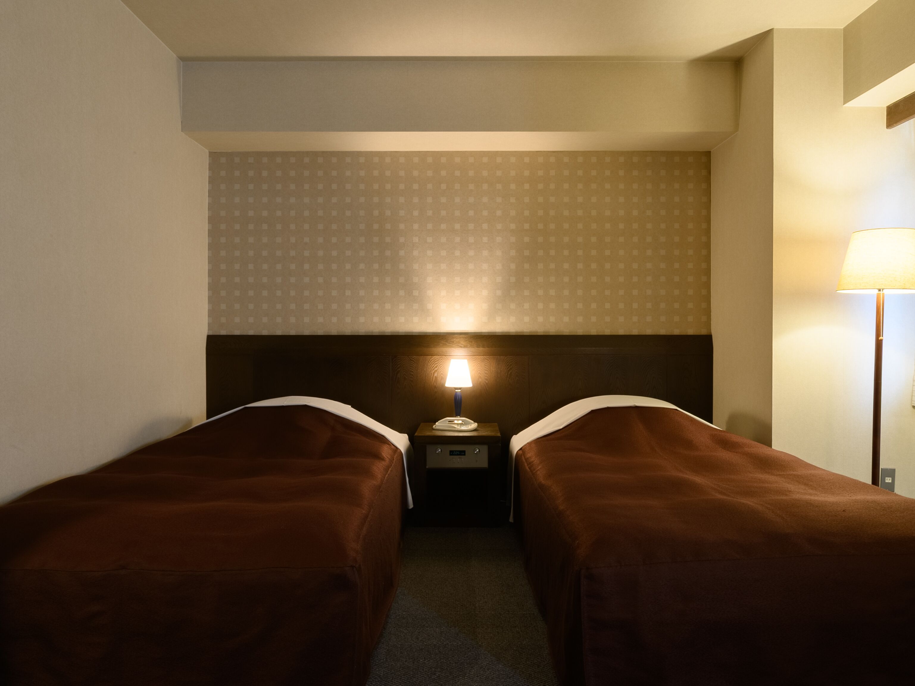 Quarto de hotel com duas camas de solteiro, mesa de cabeceira com abajur e parede estampada.