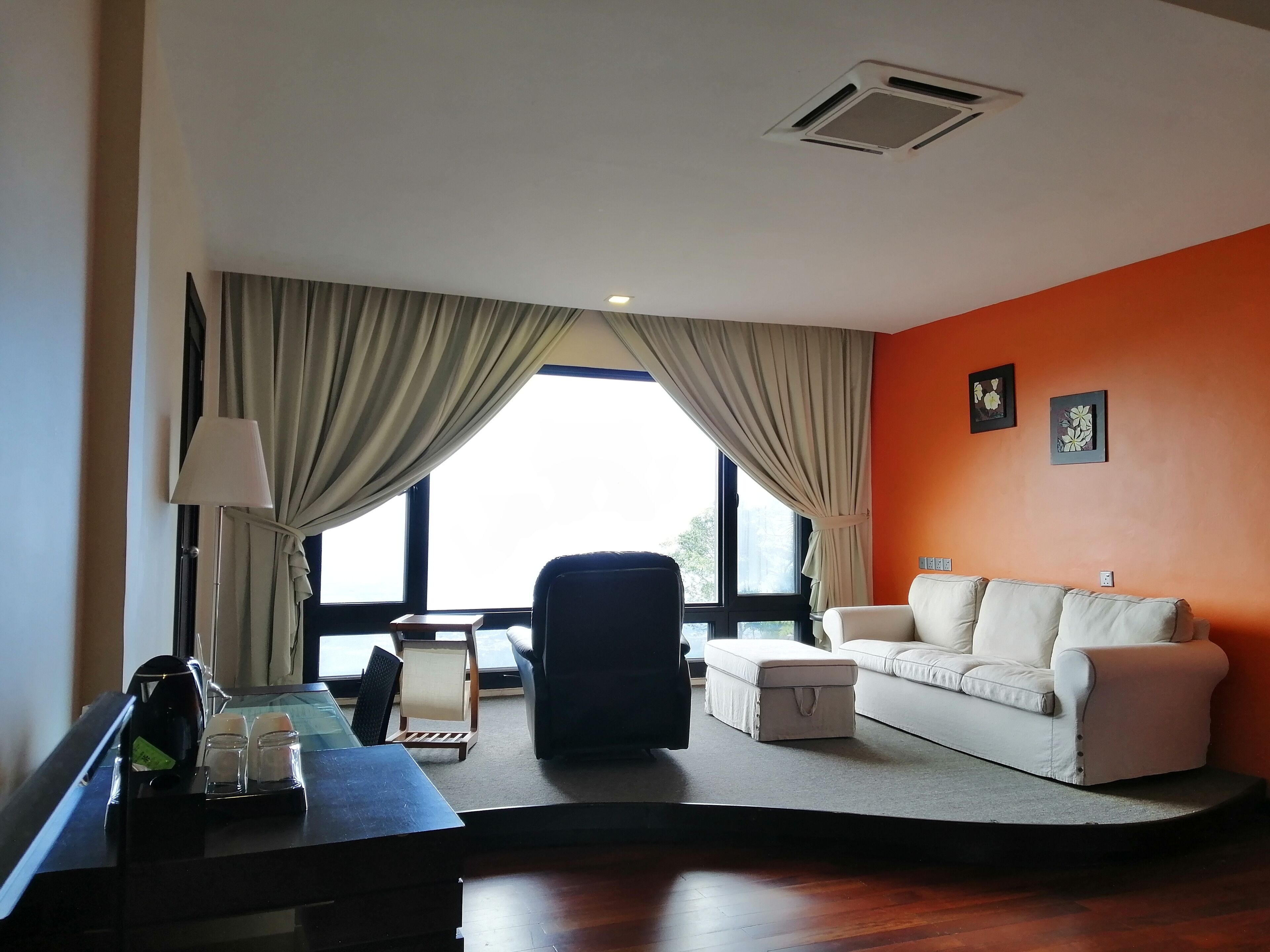 Quarto de hotel com escrivaninha, cadeira, sofá branco e uma janela grande com cortinas.