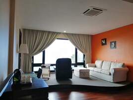 Quarto de hotel com escrivaninha, cadeira, sofá branco e uma janela grande com cortinas.
