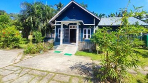 Uma casa de dois andares com telhado azul e detalhes brancos, cercada por vegetação exuberante e um céu azul límpido.