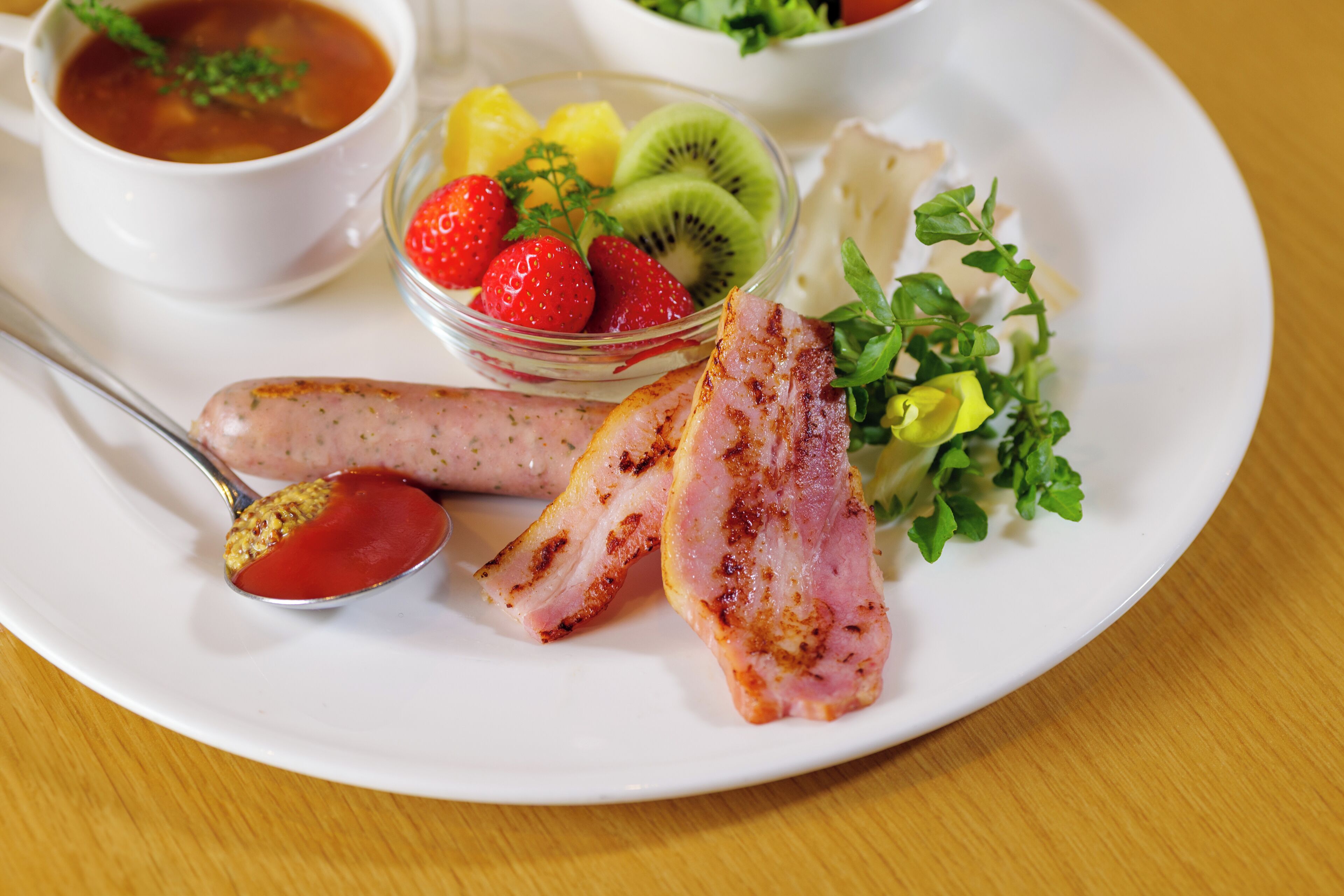 Um prato com alimentos, incluindo salsicha, bacon, morangos, kiwi e uma pequena tigela de sopa.