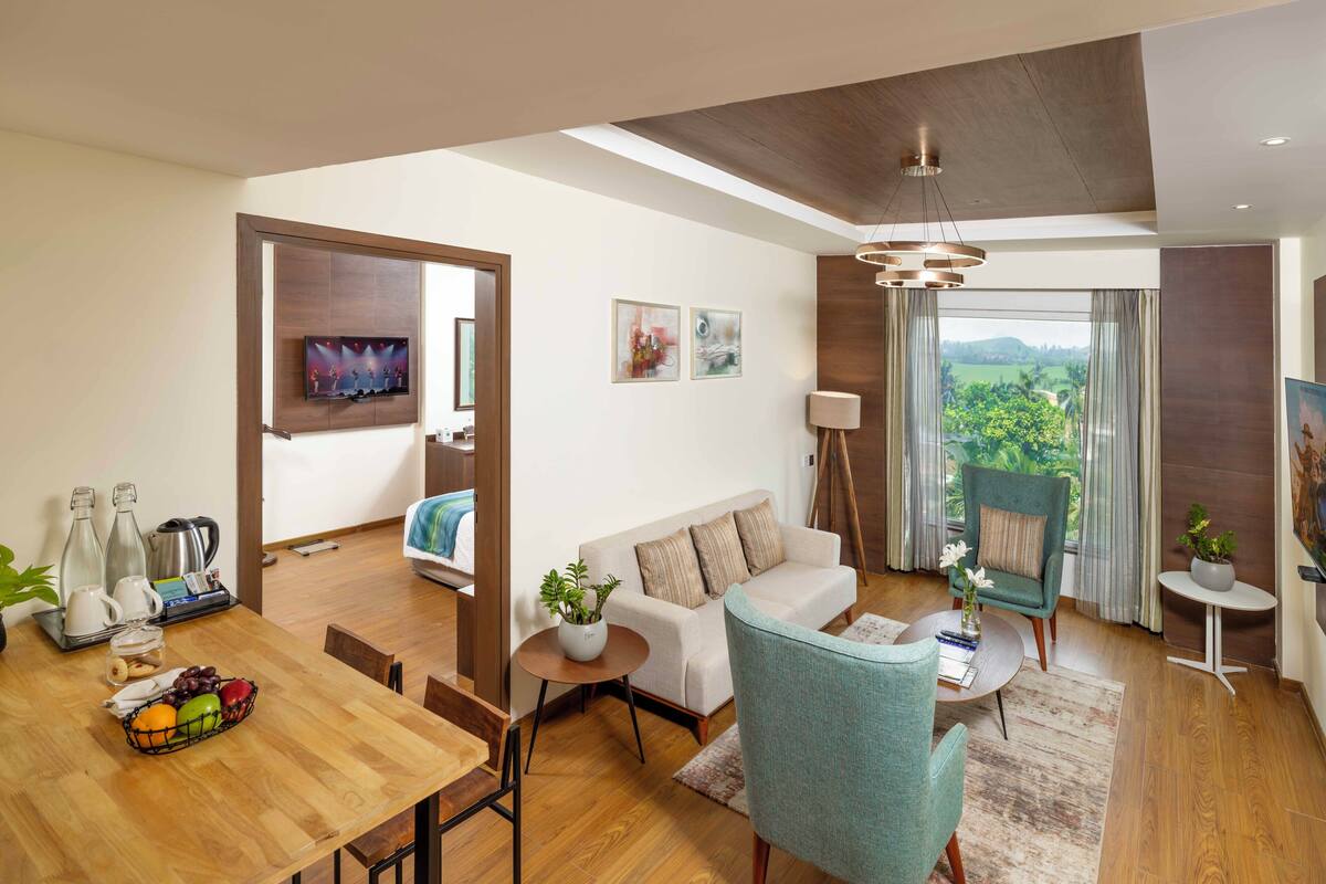 Uma sala de estar moderna com sofá, poltronas, uma mesa de jantar com cesta de frutas, um canto de café e vista para o verde através de grandes janelas.