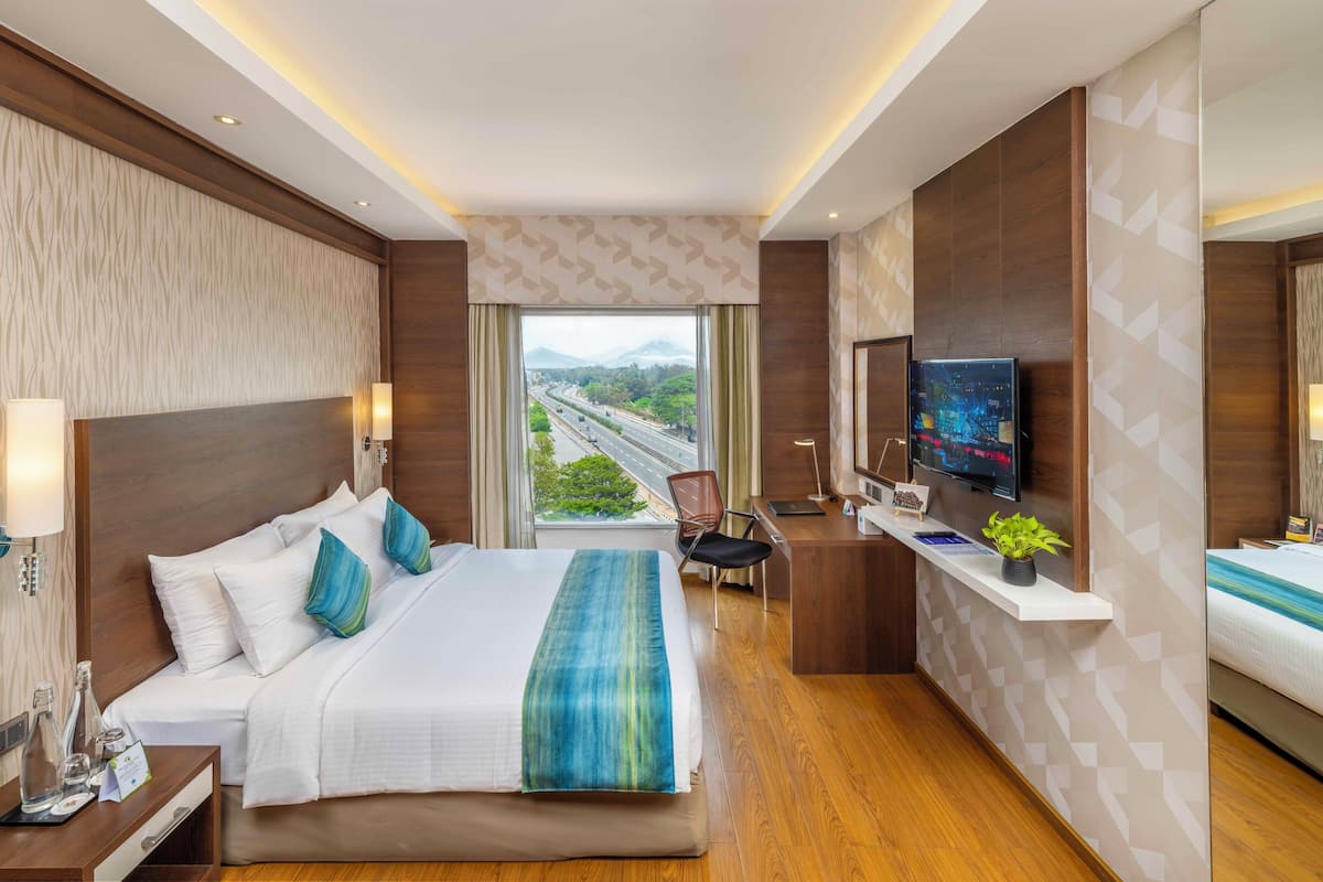 Quarto de hotel com uma cama grande, uma escrivaninha com computador, televisão e vista para uma autoestrada.