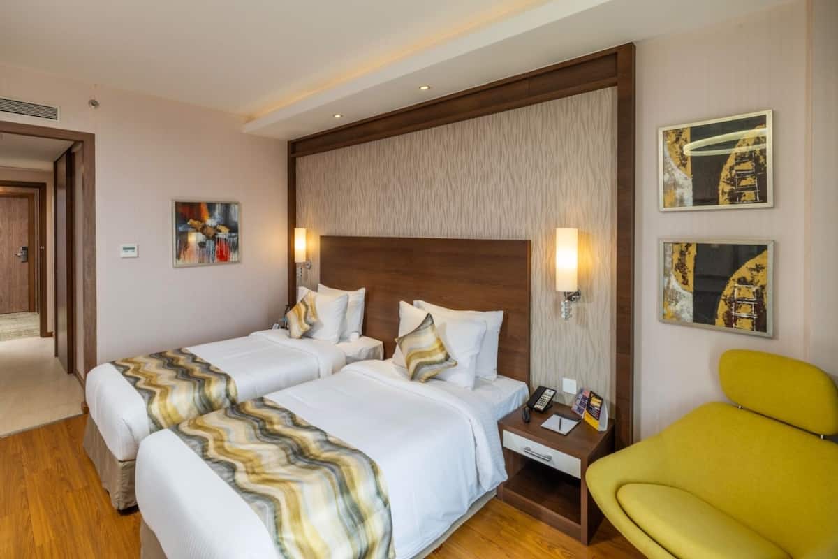 Quarto de hotel com duas camas, cabeceira de madeira, mesa de cabeceira com abajur, uma cadeira e obras de arte nas paredes.
