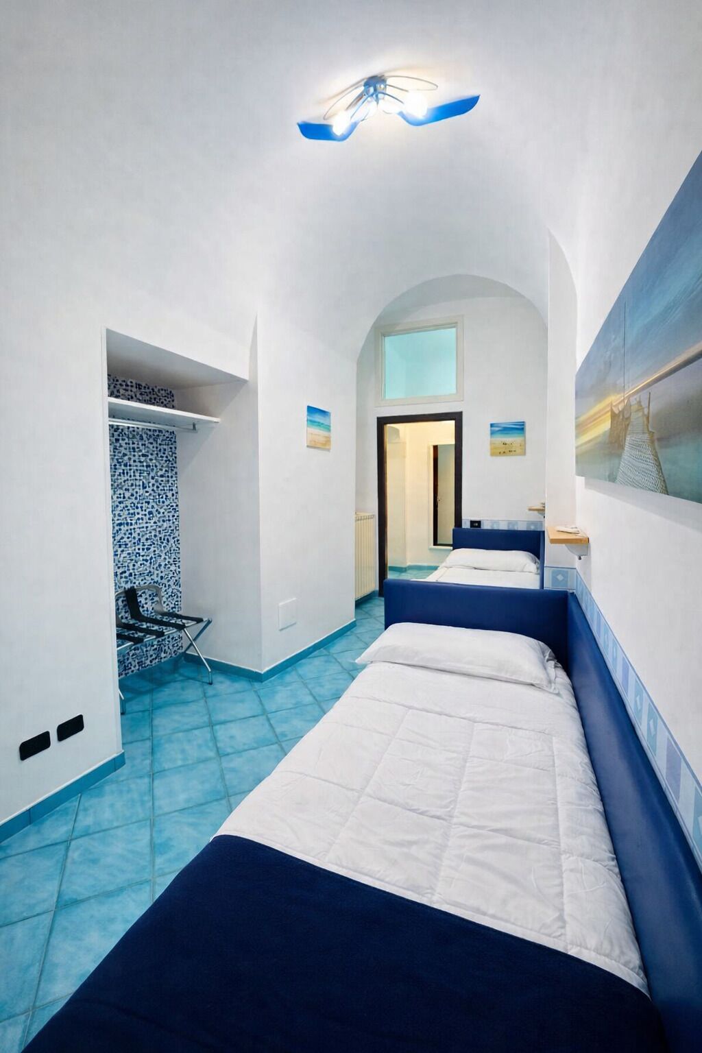 Um quarto com cama, uma cadeira, uma mesinha, um ventilador de teto e um quadro na parede.