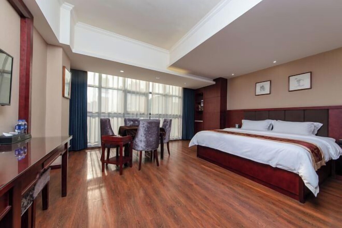 Quarto de hotel com uma cama grande, uma escrivaninha e uma cadeira. Há uma janela com cortinas e vista para o exterior.