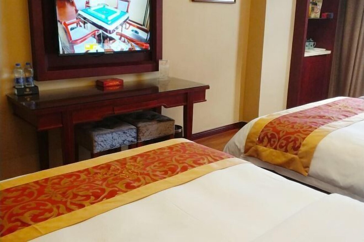 Quarto de hotel com duas camas, uma televisão sobre um suporte de madeira, uma mesinha com um objeto vermelho e uma estante de livros.