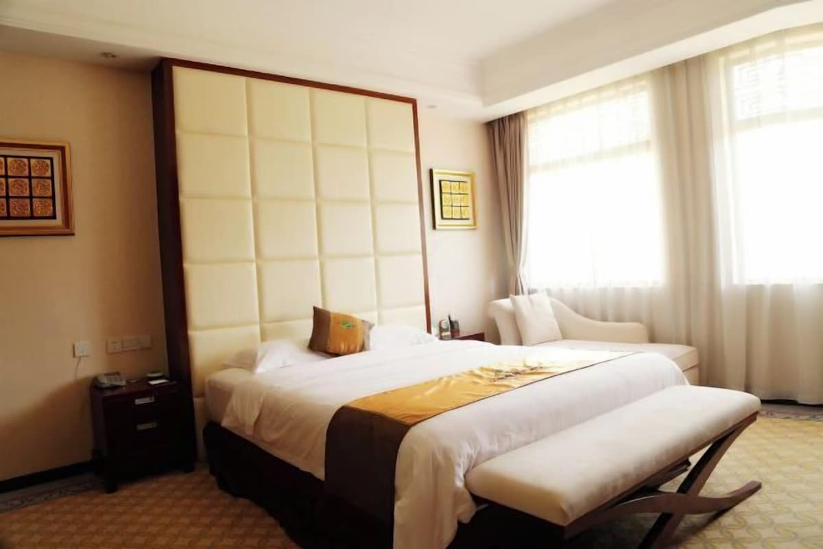 Quarto de hotel com cama grande, mesa de cabeceira, cadeira e janela com cortinas.
