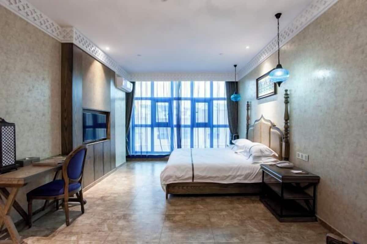 Quarto com cama, escrivaninha, cadeira e uma grande janela com cortinas.