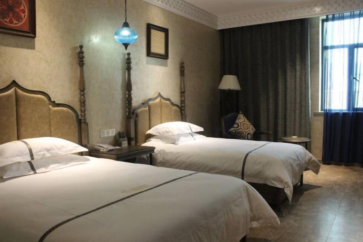 Quarto de hotel com duas camas, uma mesa de cabeceira, luminária, cadeira e janela com cortinas.