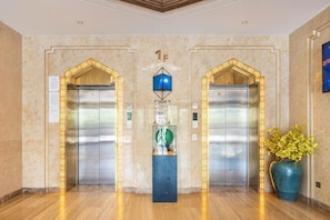 Um hall com dois elevadores, um vaso azul com flores amarelas e um relógio na parede.