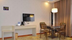 Quarto de hotel com televisão de tela plana na parede, uma escrivaninha com cafeteira, mesa de jantar com cadeiras e uma janela grande com cortinas.