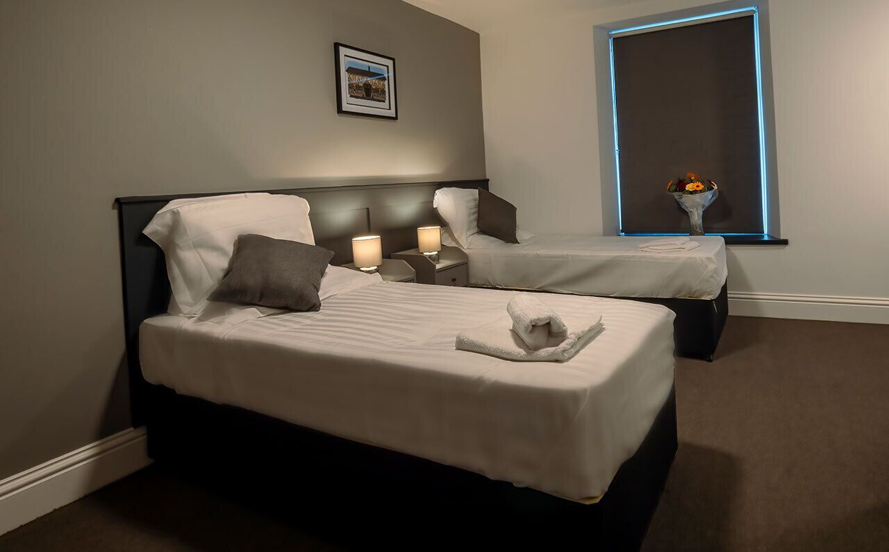 Quarto de hotel com duas camas, cada uma com um travesseiro, uma mesinha de cabeceira com abajur, e uma janela com luz azul.